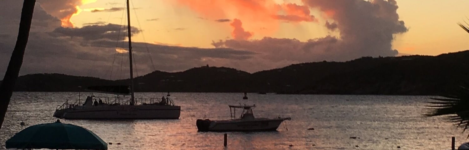 Um pôr do sol sobre a água, com barcos, palmeiras e área de piscina.