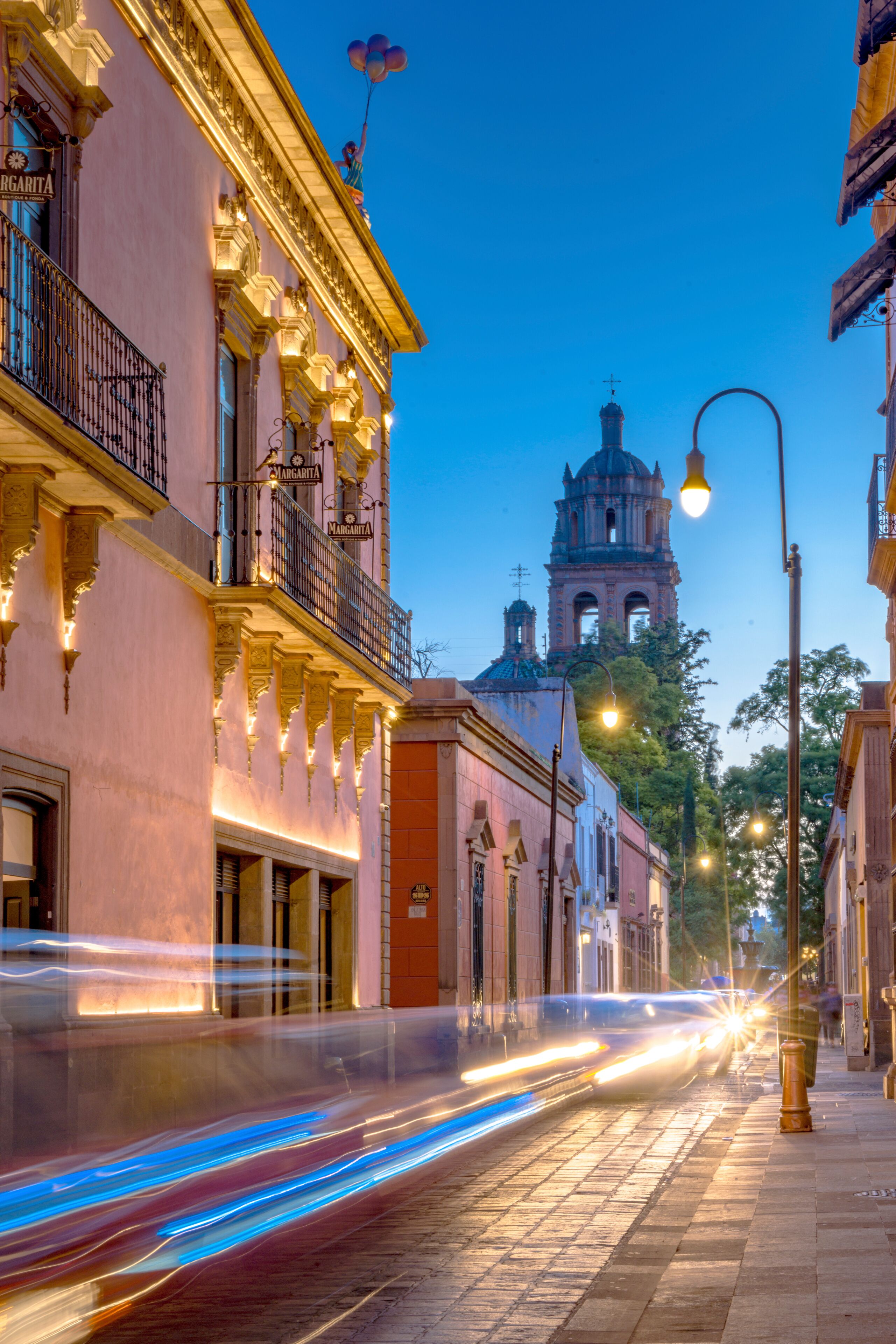 Uma rua histórica com edifícios ornamentados, uma torre de sino imponente e rastros de luz deixados por veículos.