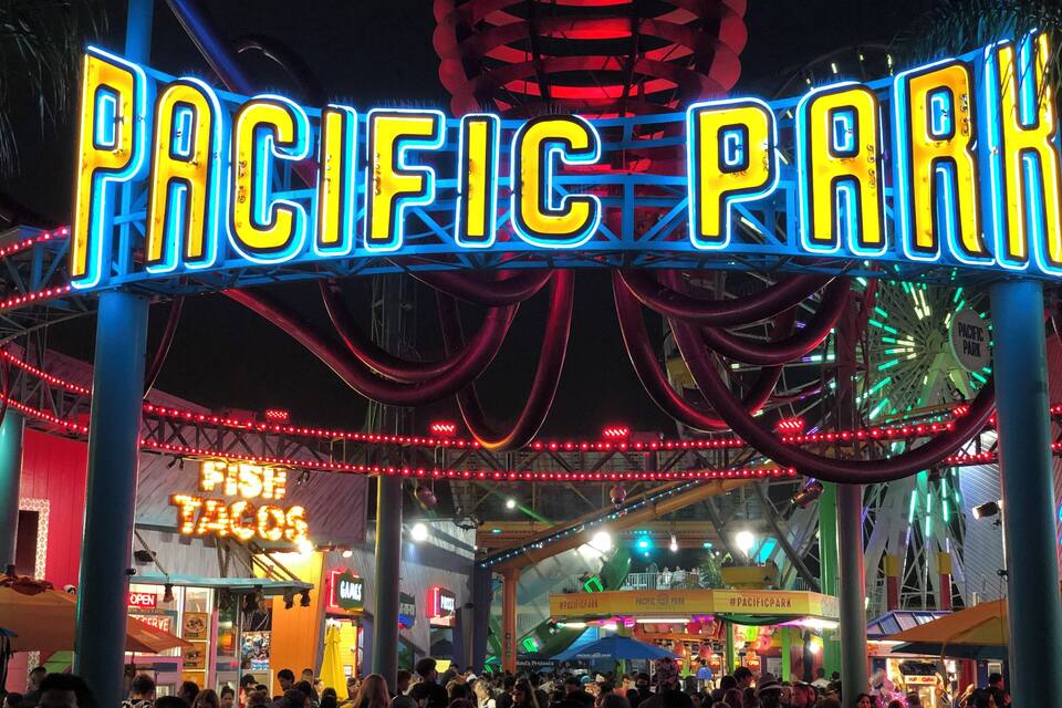 Uma entrada de parque de diversões bem iluminada, com letreiros de neon, incluindo "PACIFIC PARK" e "FAST TACO'S".