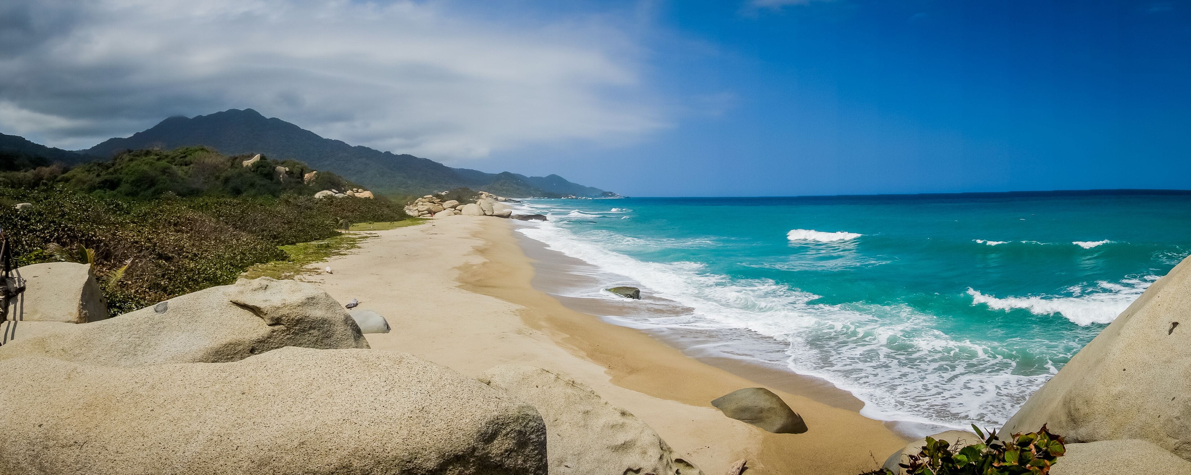 Uma praia com ondas, montanhas ao fundo e céu claro.