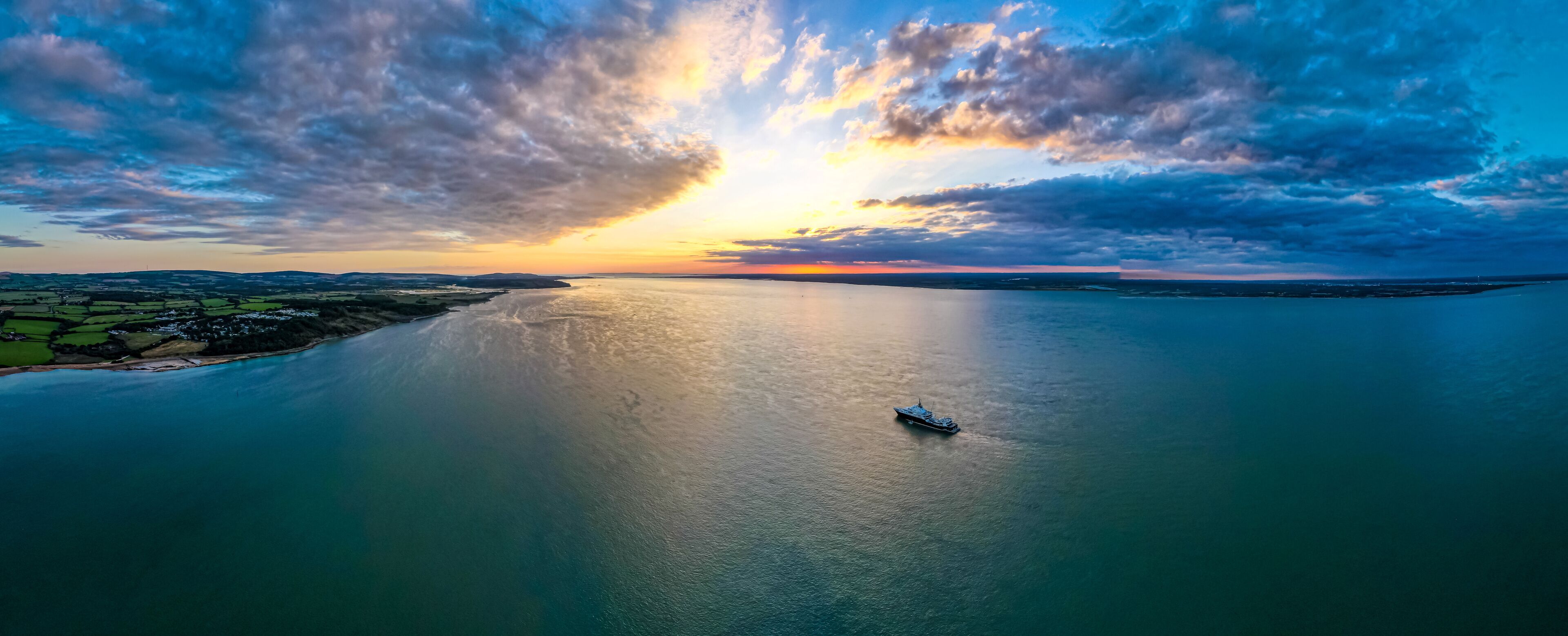 Um pôr do sol sobre um mar calmo, com um barco, terra ao longe e um céu nublado.