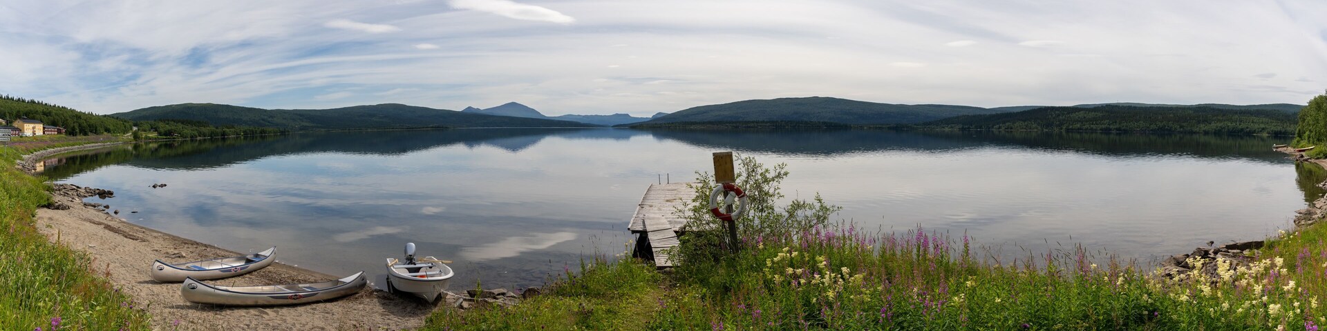 Um lago calmo com barcos na margem, rodeado por montanhas e vegetação.