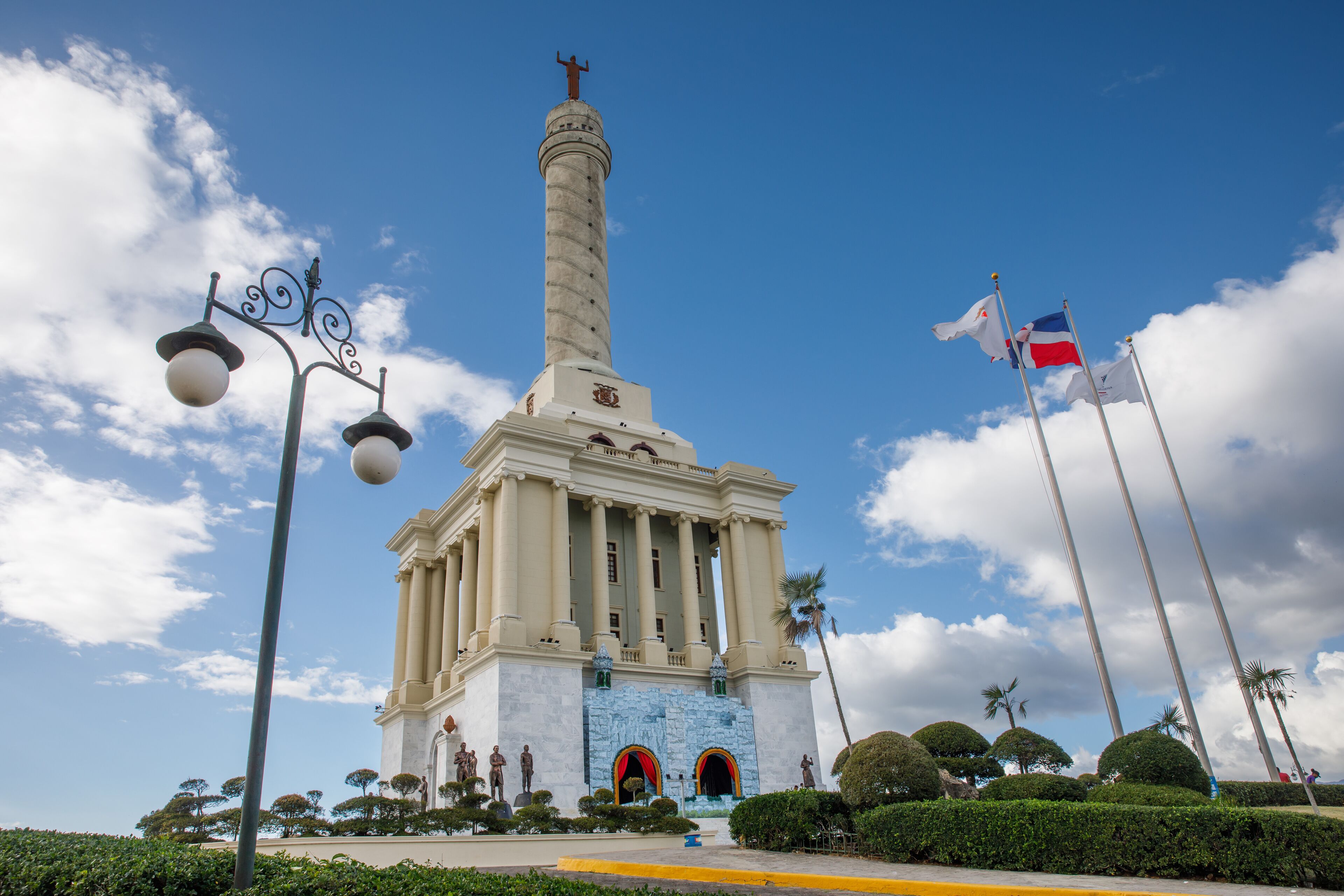 Um monumento com uma coluna alta e uma cruz no topo, cercado por estátuas e bandeiras.