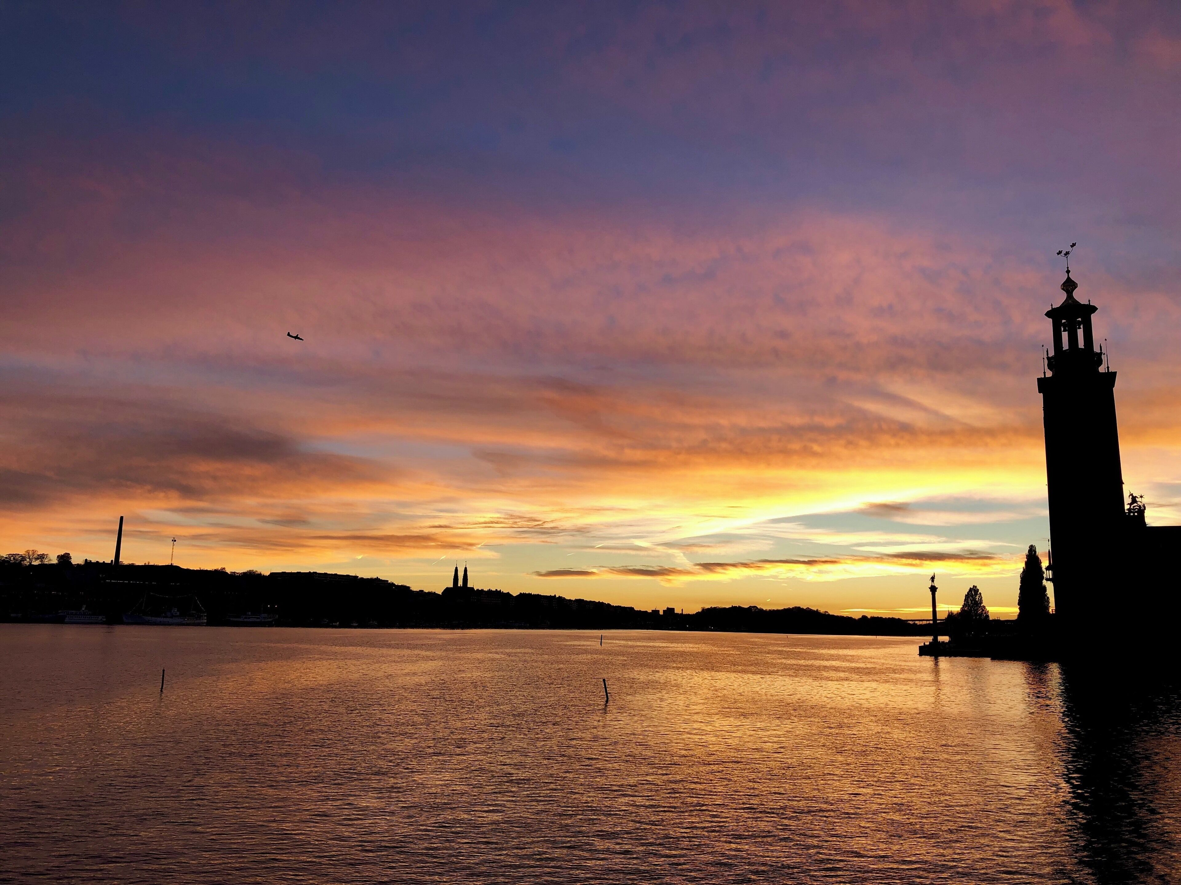 Um pôr do sol sobre a água, com uma torre e duas torres pontiagudas recortadas no céu.