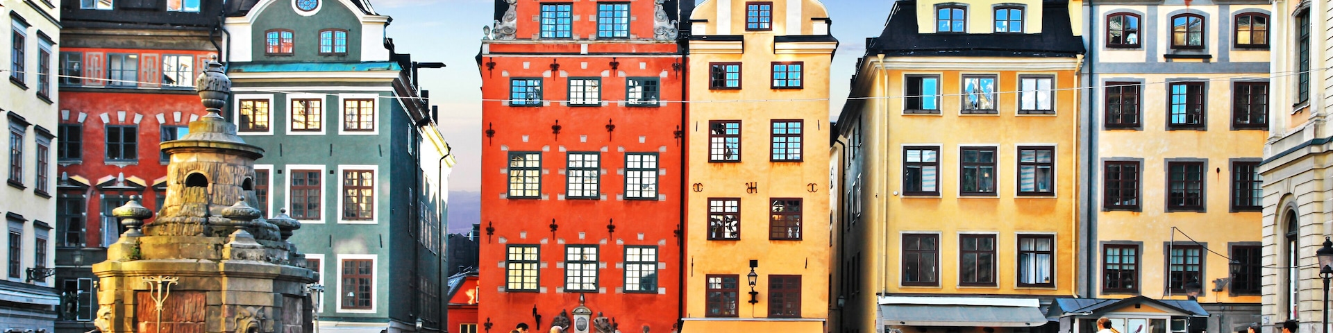 Uma praça de paralelepípedos com edifícios coloridos, uma fonte e bancos.