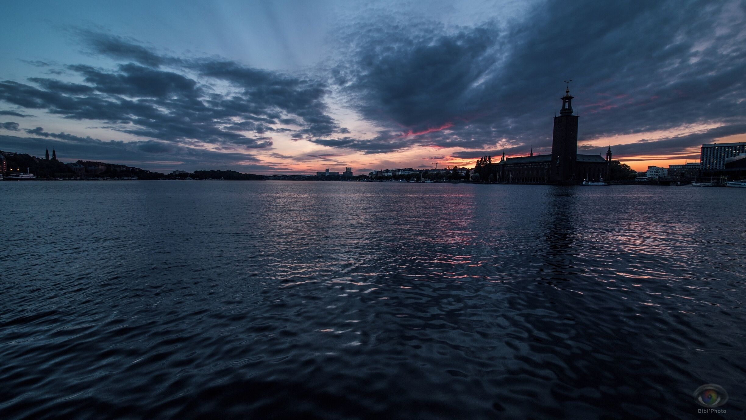 Um pôr do sol sobre a água, com uma torre imponente e edifícios ao fundo.