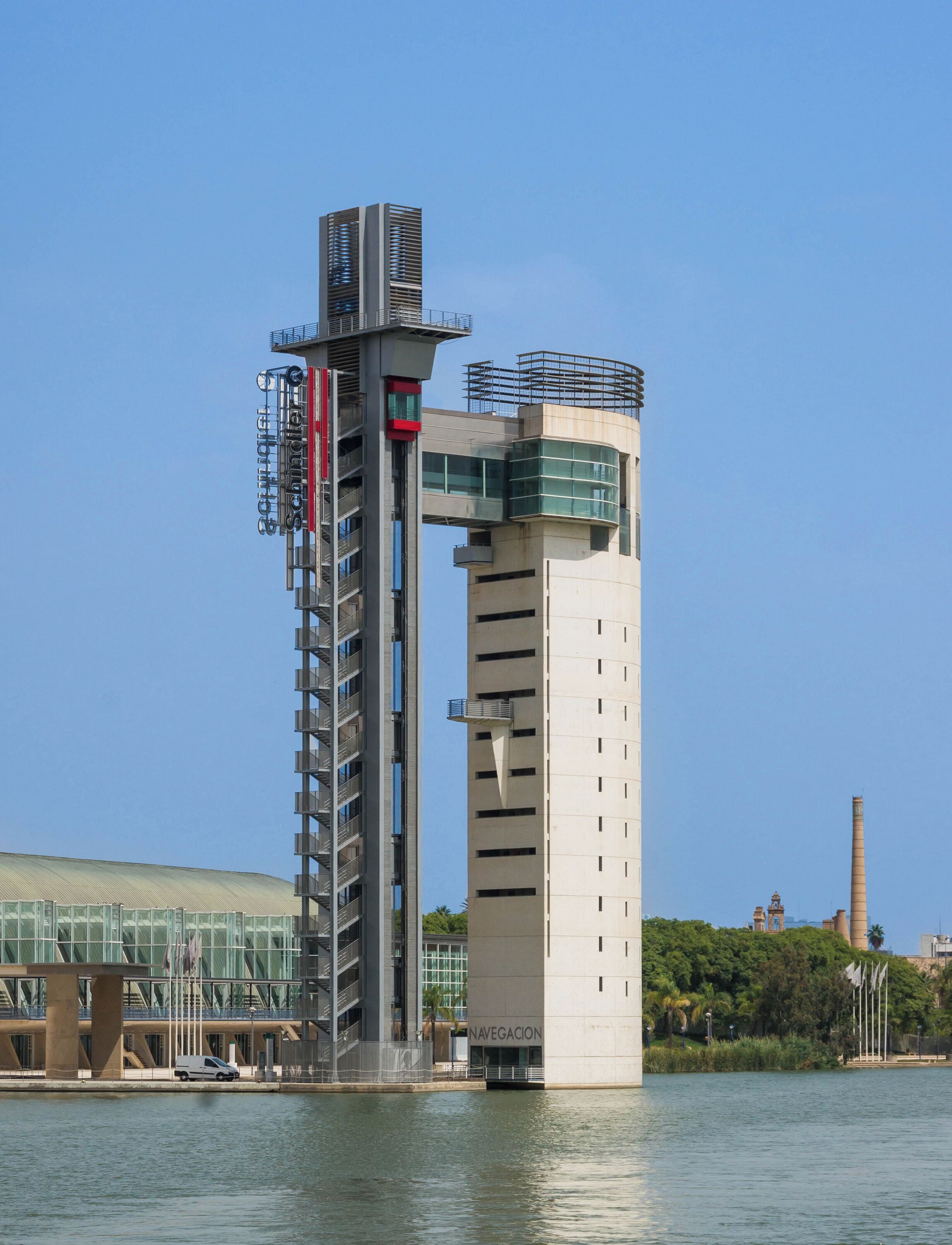 Uma torre moderna com fachada de vidro e um detalhe vermelho, localizada perto de um corpo d'água.