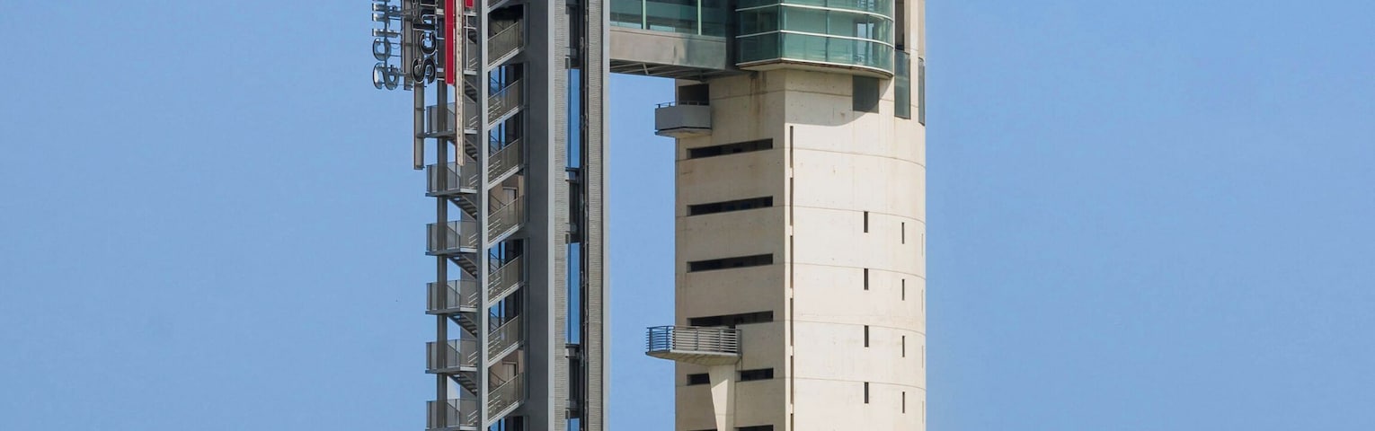Uma torre moderna com fachada de vidro e um detalhe vermelho, localizada perto de um corpo d'água.