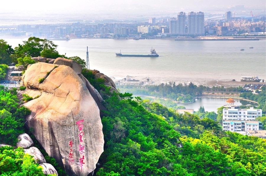 Uma grande rocha com caracteres chineses, um rio, o horizonte de uma cidade e uma ponte ao longe.