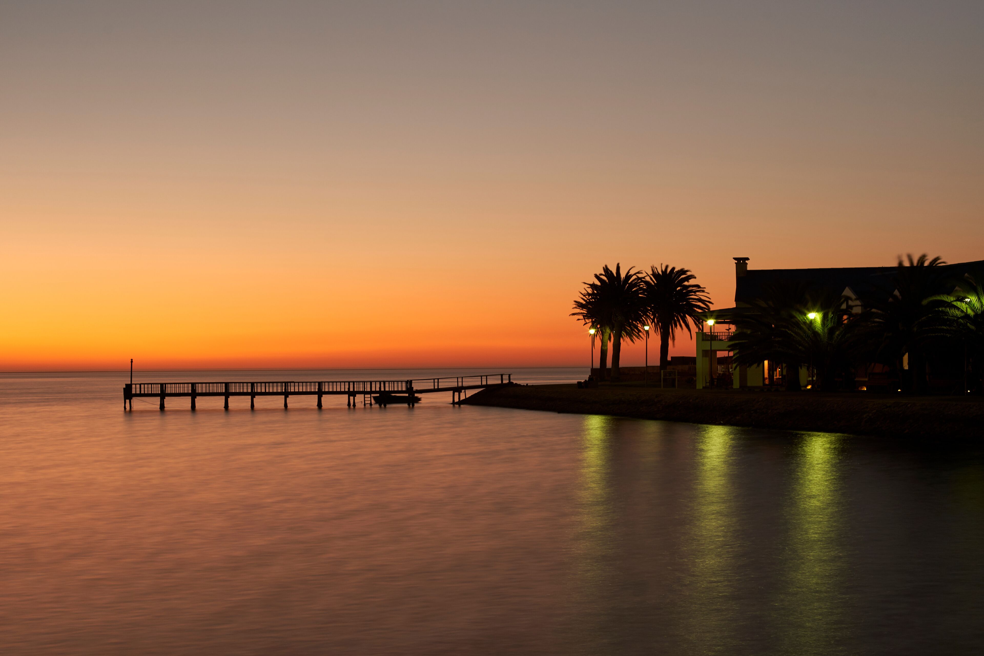 Um pôr do sol sobre um mar calmo, com um píer que se estende na água, palmeiras e um edifício iluminado à direita.