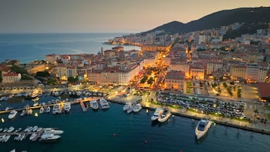 Paisagem costeira ao entardecer com um porto cheio de barcos, edifícios históricos e um horizonte com colinas.