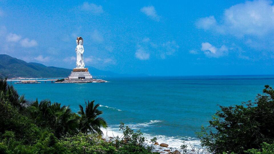 Uma estátua numa pequena ilha rodeada por águas azul-turquesa e colinas verdes.