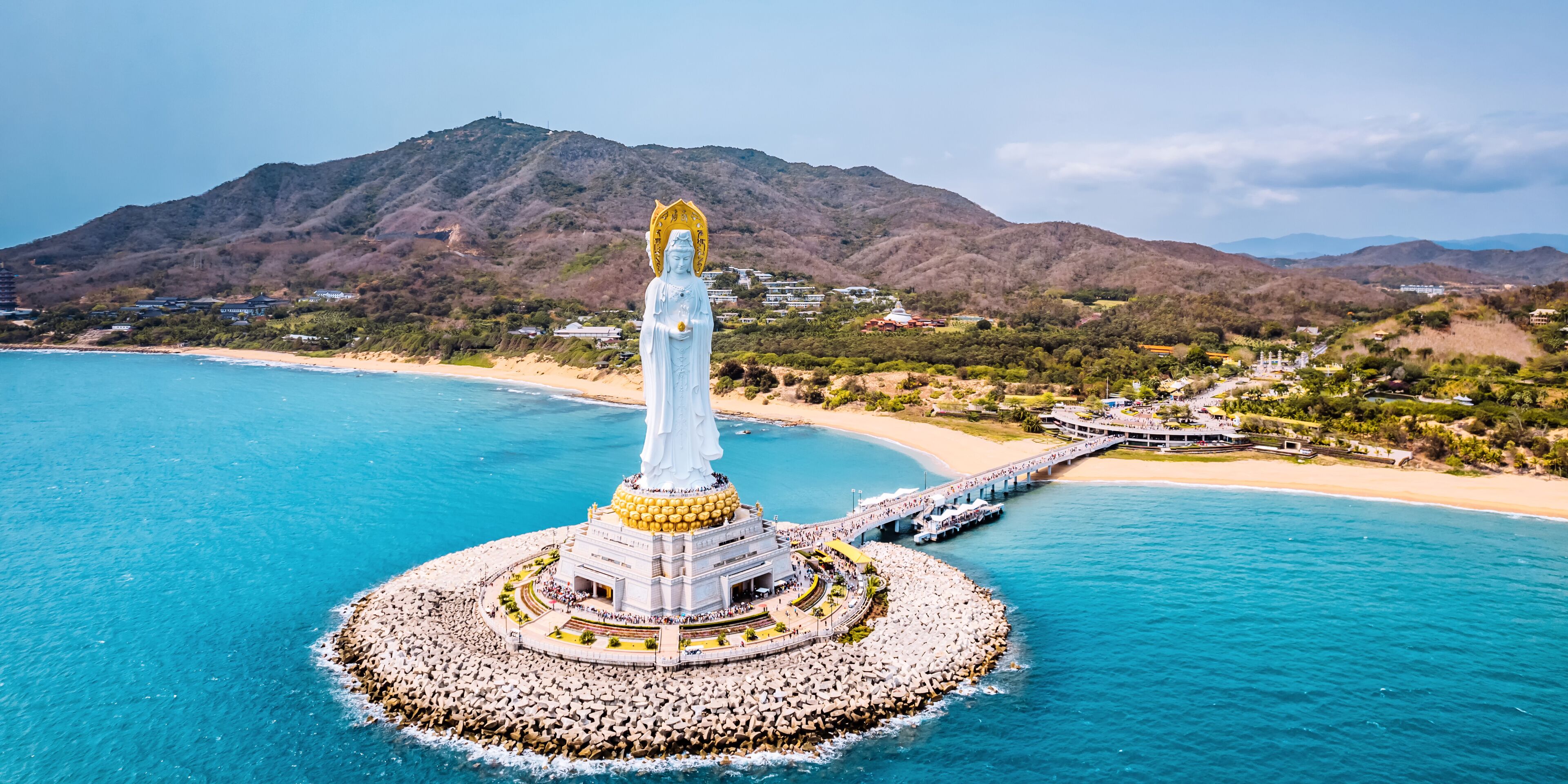 Uma estátua branca sobre um pedestal de pedra circular, rodeada por água, com uma praia e montanhas ao fundo.