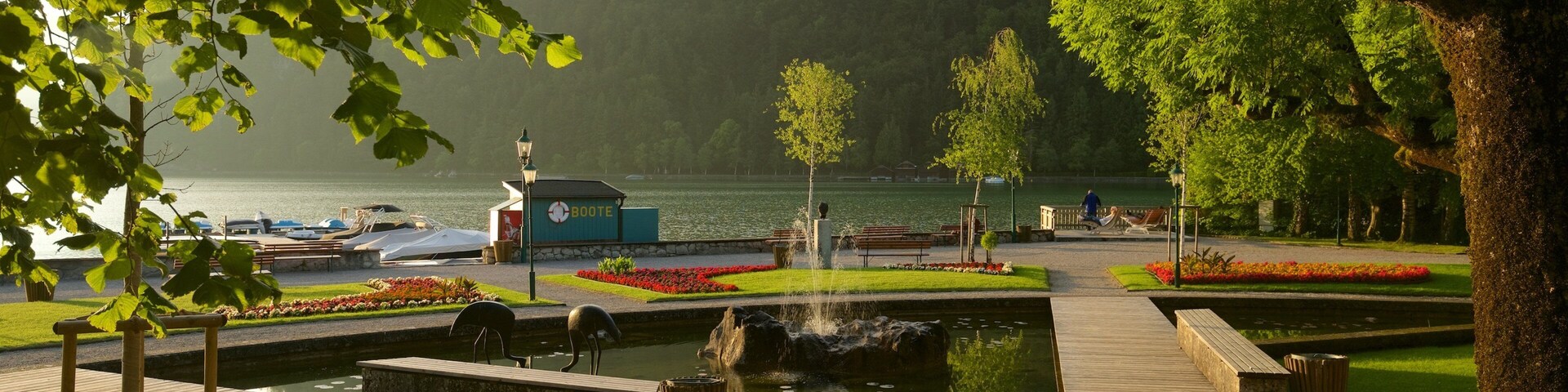 Um parque à beira do lago com um cais de madeira, uma fonte, bancos e um barco abrigo com a inscrição 'BOOTE'.