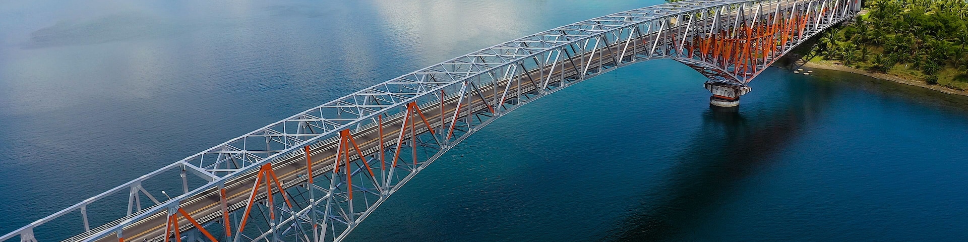 Uma extensa ponte de aço atravessa um rio largo, rodeada por vegetação exuberante e colinas distantes.