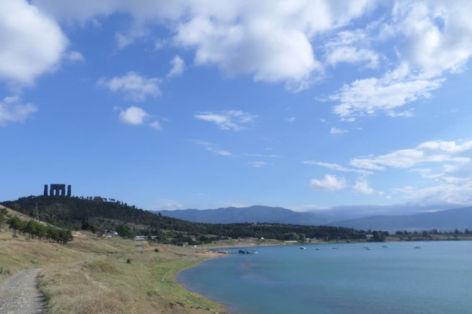 Paisagem costeira com uma colina com ruínas, céu claro e mar calmo com barcos.
