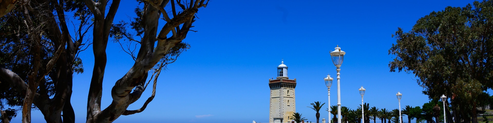 Um calçadão costeiro com um farol, árvores e um céu azul límpido.