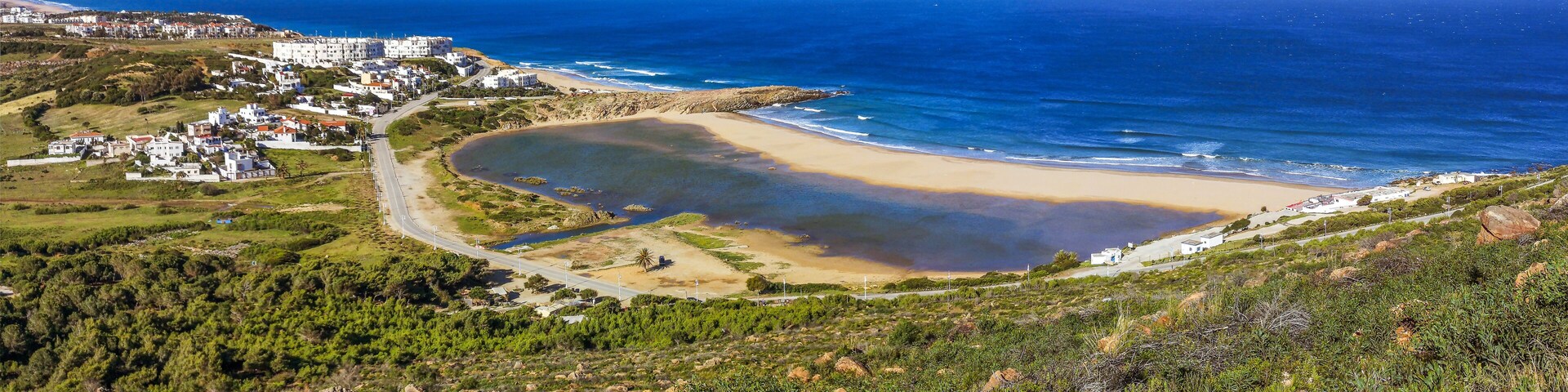 Paisagem costeira com praia, afloramentos rochosos e algumas construções espalhadas.