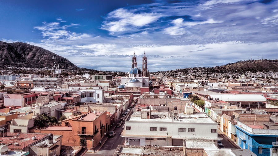 Paisagem urbana com uma igreja imponente e duas torres, cercada por edifícios próximos e um horizonte montanhoso.