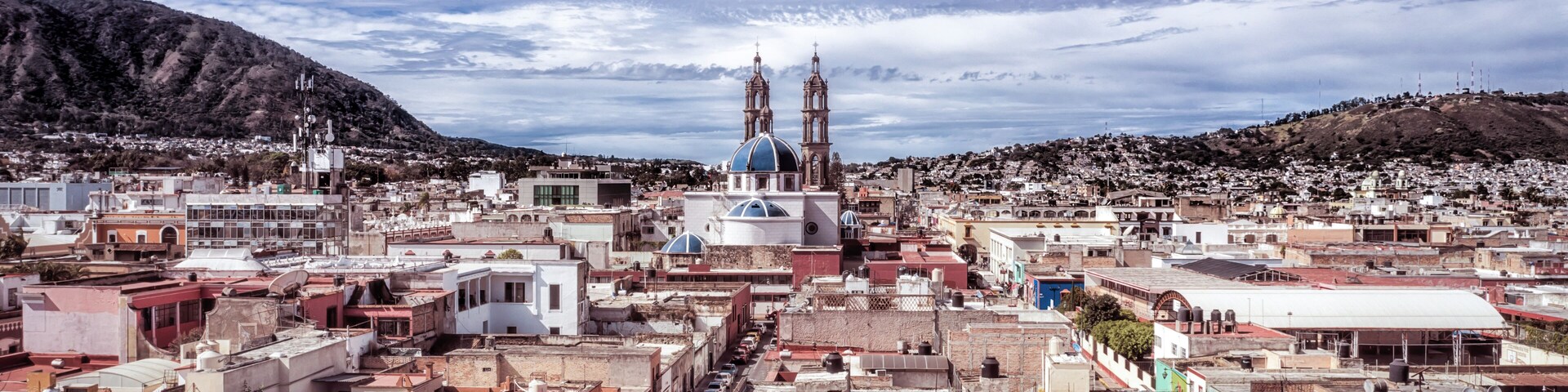 Paisagem urbana com uma igreja imponente e duas torres, cercada por edifícios próximos e um horizonte montanhoso.
