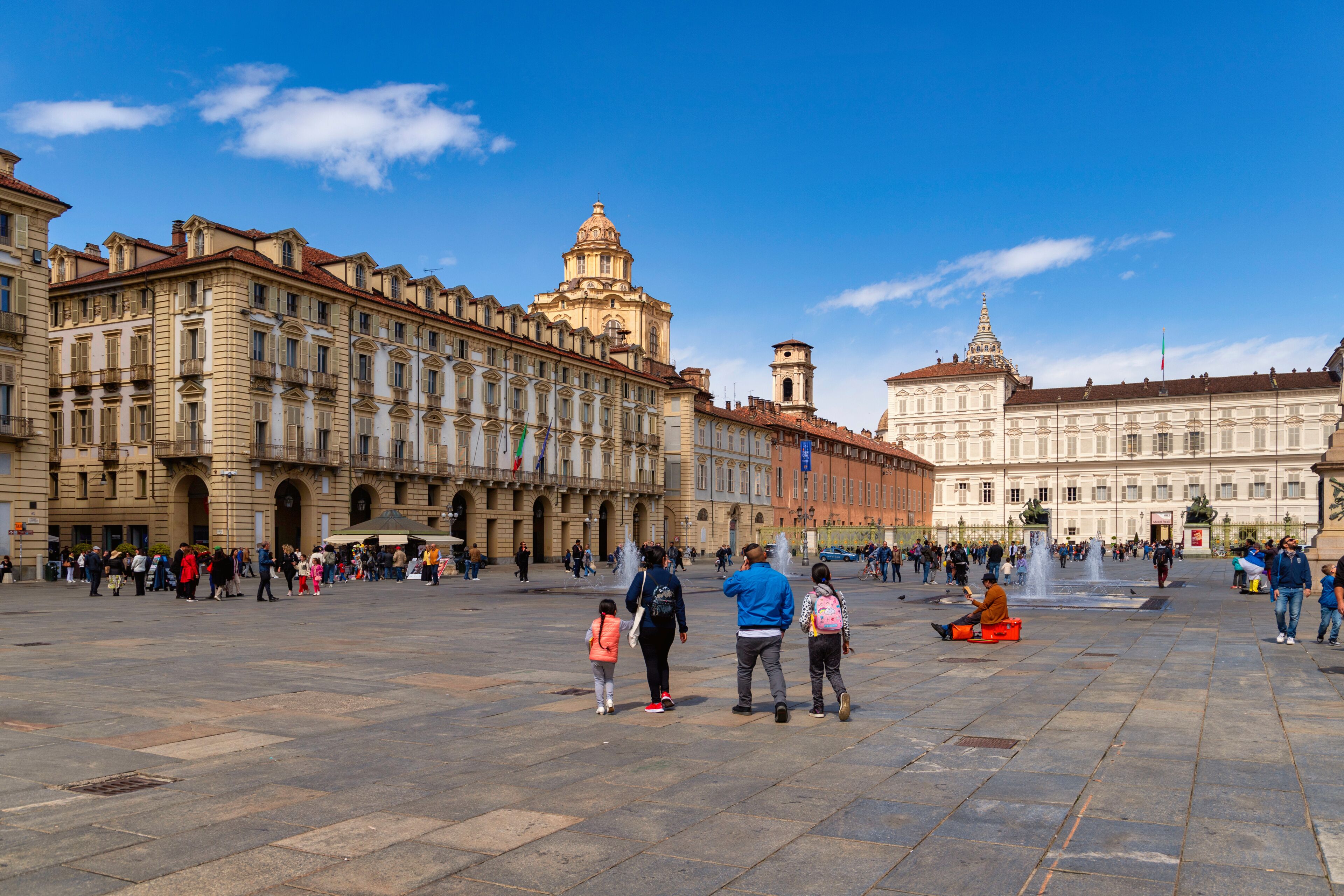 Uma praça histórica com edifícios elaborados, uma fonte e pessoas caminhando.