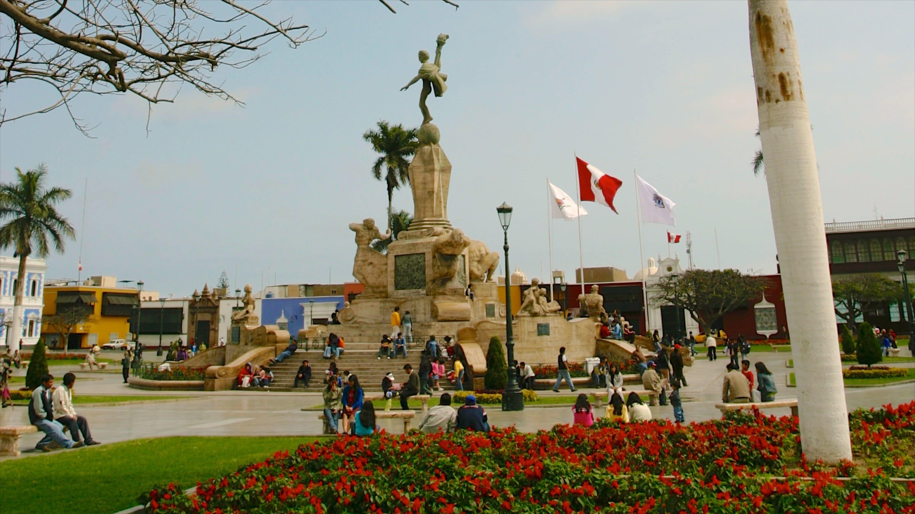 Uma praça pública com um monumento central, palmeiras e flores vermelhas.