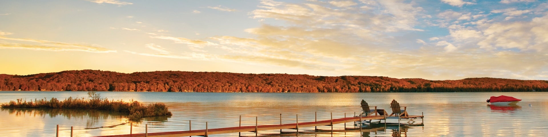 Um lago calmo com um cais de madeira que se estende pela água, rodeado por árvores com folhagem outonal e um céu limpo.