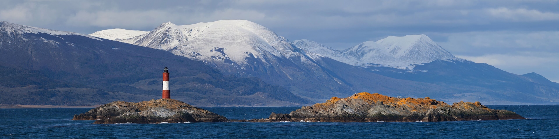 Um farol numa ilha rochosa com montanhas cobertas de neve ao fundo.