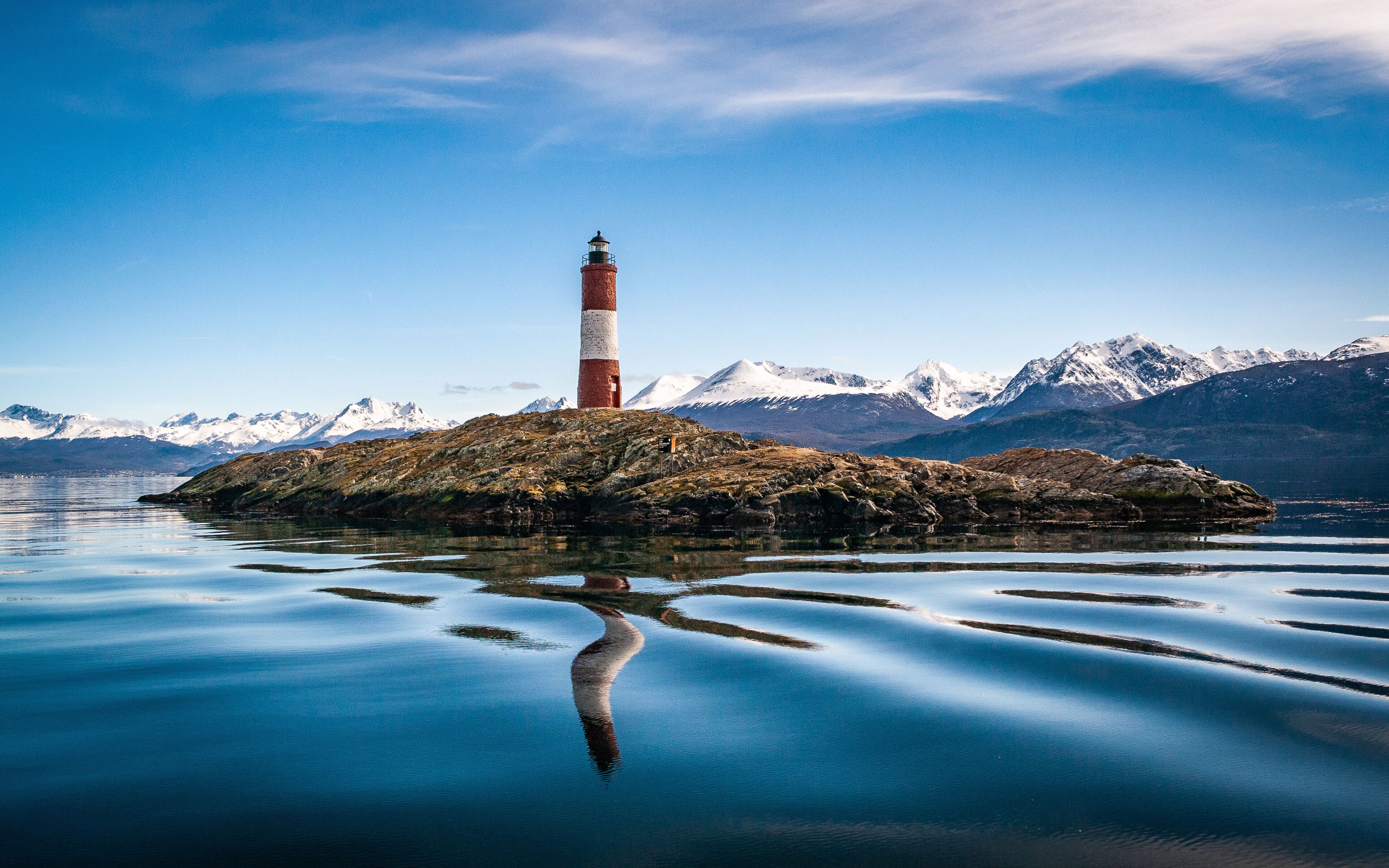 Um farol numa ilha rochosa, com montanhas cobertas de neve ao fundo.