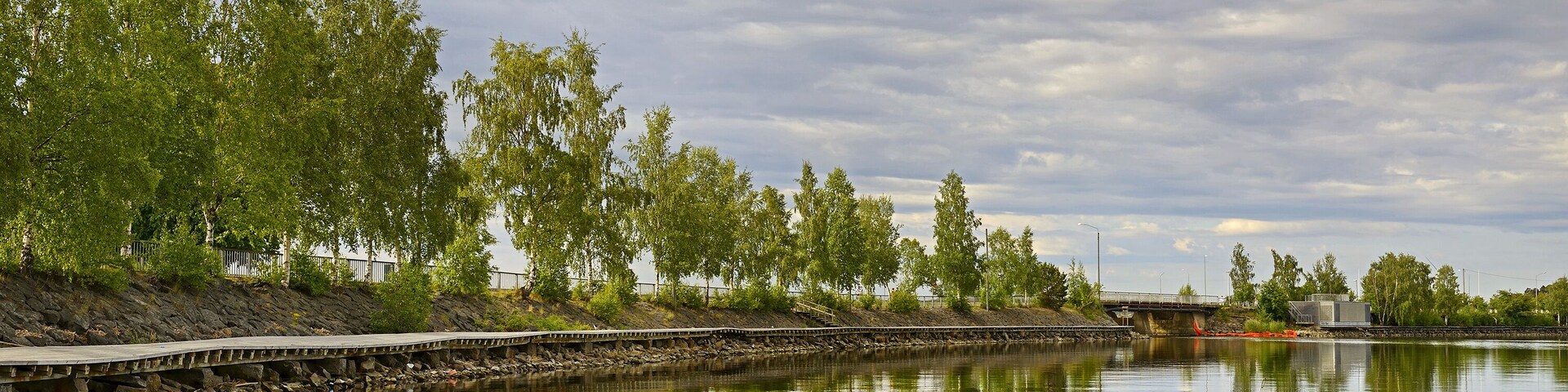 Um rio calmo com um talude de pedra, árvores verdes e céu nublado.