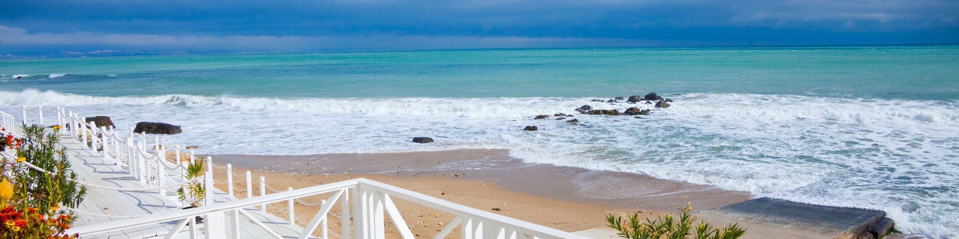 Um calçadão de madeira leva a uma praia com águas azul-turquesa e céu nublado.