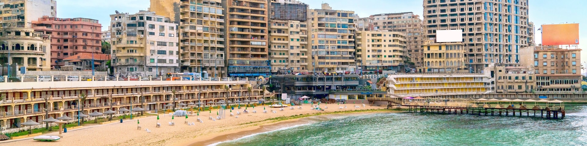 Paisagem costeira urbana com uma praia de areia, um píer que se estende sobre a água e diversos edifícios altos.