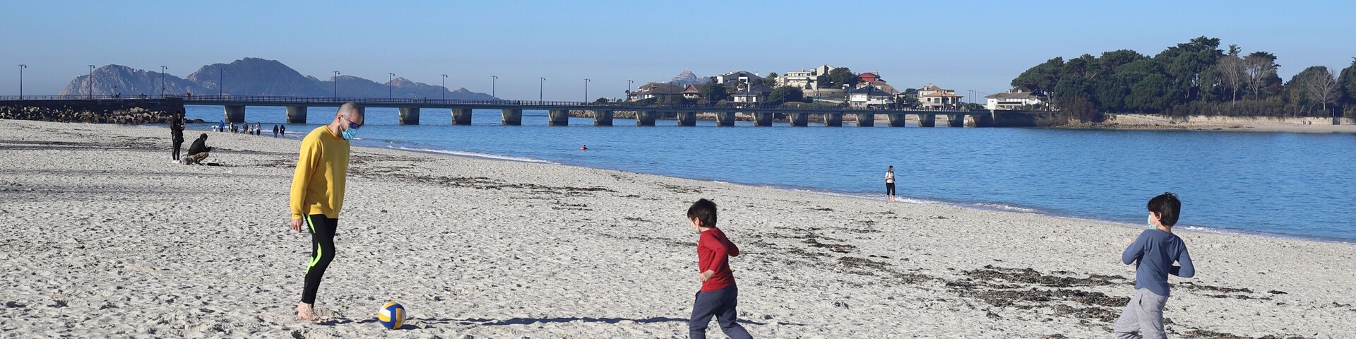 Uma praia arenosa com uma ponte ao fundo e um céu azul límpido.