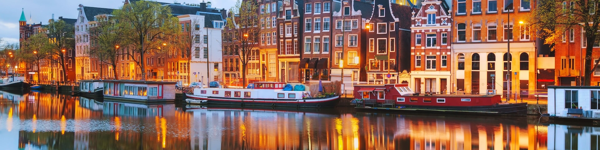 Um canal com edifícios históricos refletidos na água, um barco atracado à direita e uma rua arborizada.