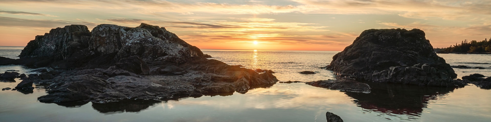 Pôr do sol sobre um mar calmo, com duas formações rochosas em destaque ao primeiro plano.
