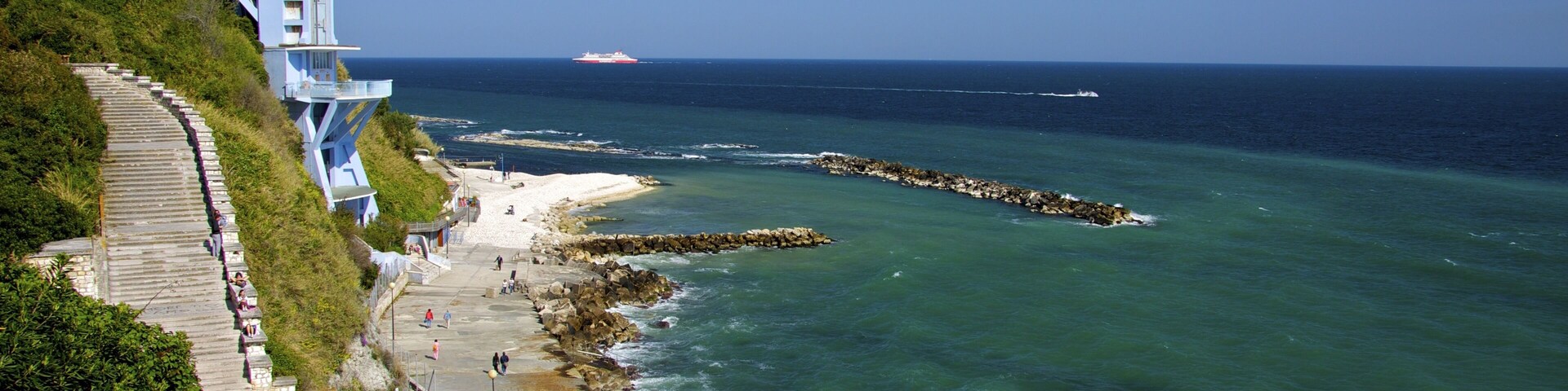 Uma escadaria costeira leva a um farol, ondas do oceano e um navio ao longe.