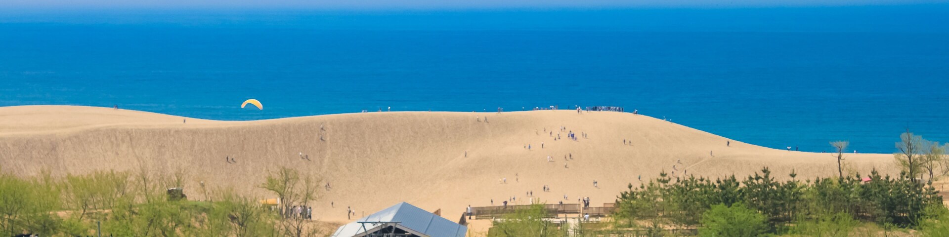 Paisagem costeira com uma praia arenosa, uma duna e um pequeno edifício com telhado azul.