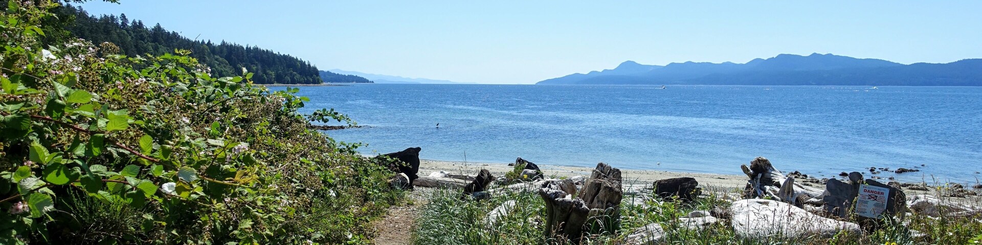 Trilho costeiro que leva a uma praia com rochas e uma vista clara do oceano e montanhas ao longe.
