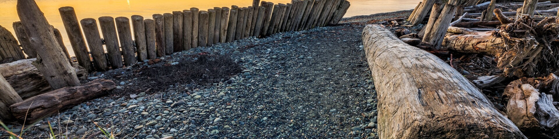 Uma praia de pedras com destroços de madeira e uma cerca rústica, com vista para um lago calmo e montanhas ao longe.
