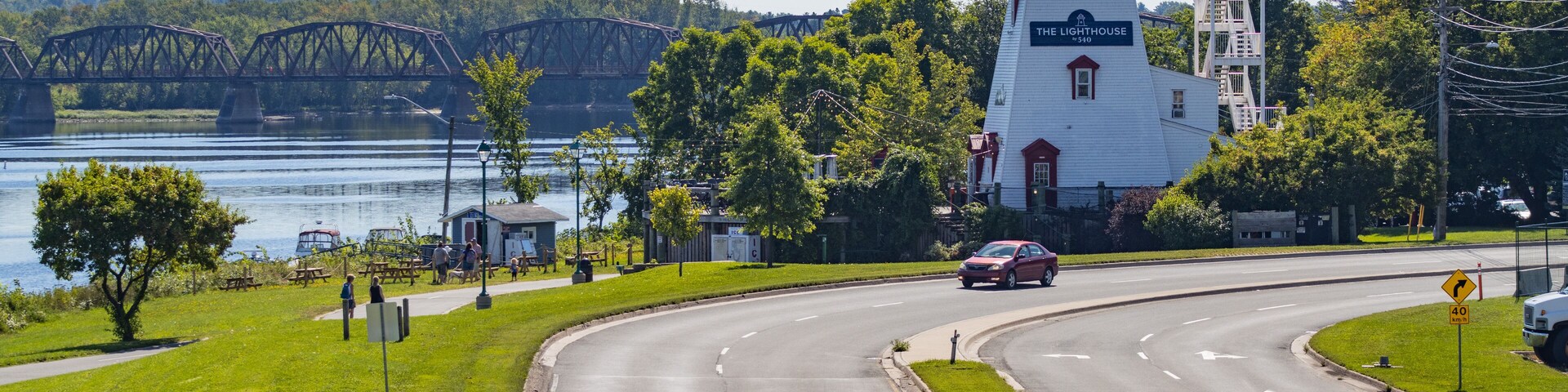 Um farol branco com teto vermelho, uma ponte ao fundo e uma estrada serpenteando ao redor do farol.