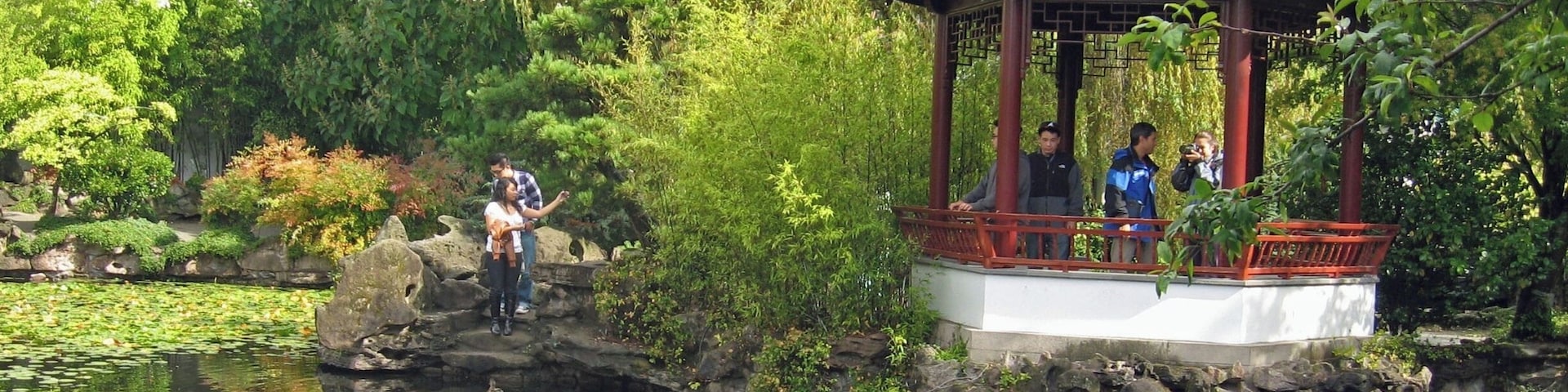 Um pavilhão chinês tradicional à beira de um lago com vegetação exuberante e uma ponte de pedra.