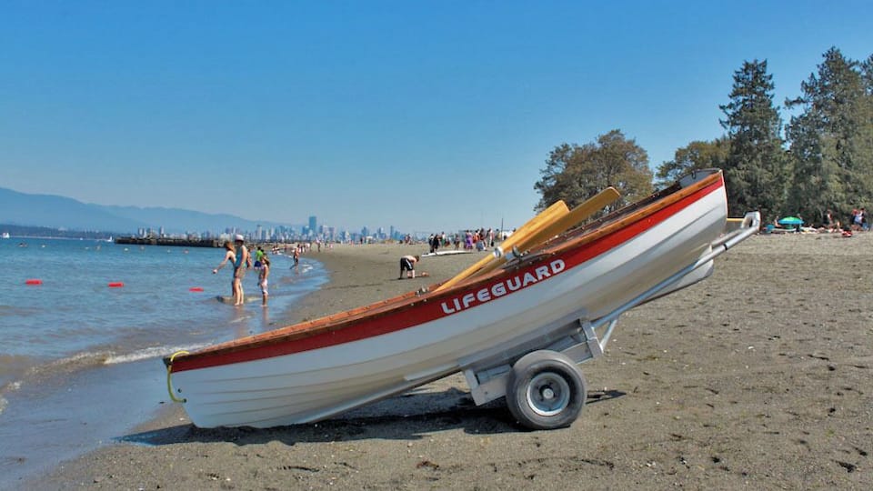 Um barco salva-vidas numa praia arenosa, com pessoas na água e um horizonte urbano ao fundo.