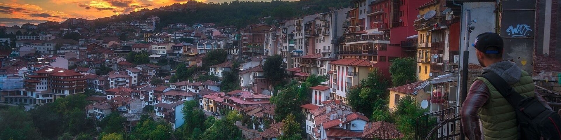 Paisagem urbana ao pôr do sol, com uma pessoa em uma varanda admirando a cidade.