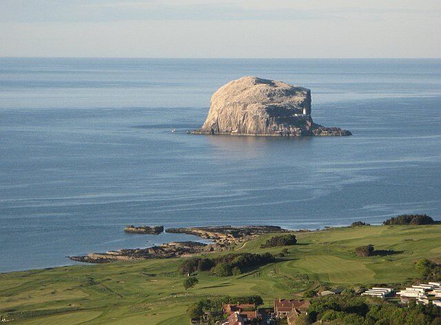 Paisagem costeira com uma formação rochosa proeminente no mar, um campo de golfe e edifícios dispersos.