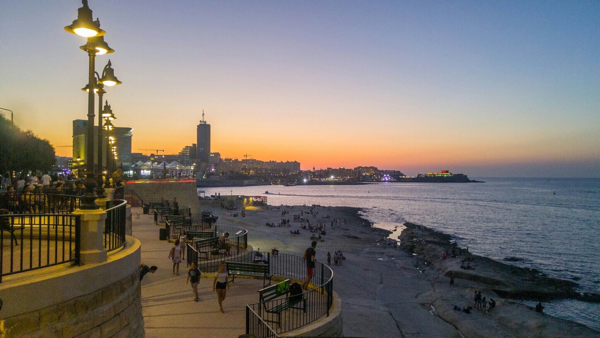 Um calçadão costeiro ao pôr do sol, com uma praia, guarda-corpo do calçadão e o horizonte da cidade ao fundo.