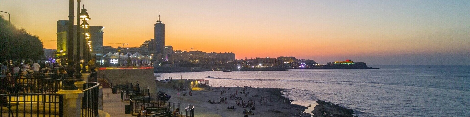 Um calçadão costeiro ao pôr do sol, com uma praia, guarda-corpo do calçadão e o horizonte da cidade ao fundo.