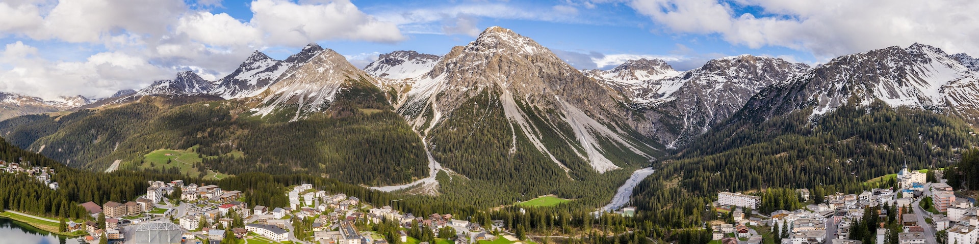 Paisagem montanhosa com uma cidade aninhada na base, um lago e picos nevados ao fundo.