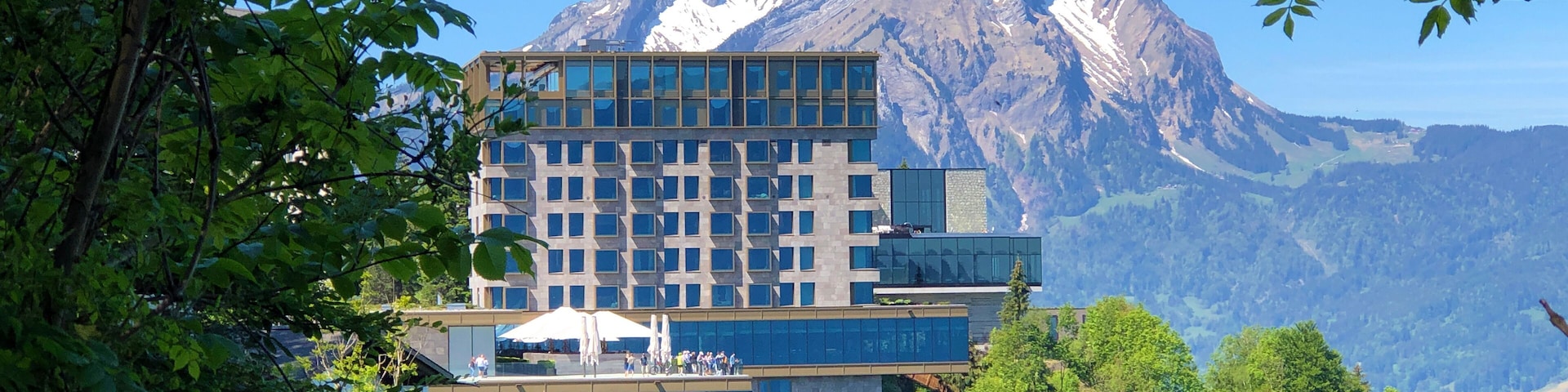 Um edifício moderno com fachada de vidro, rodeado por vegetação exuberante e uma cadeia de montanhas ao fundo.