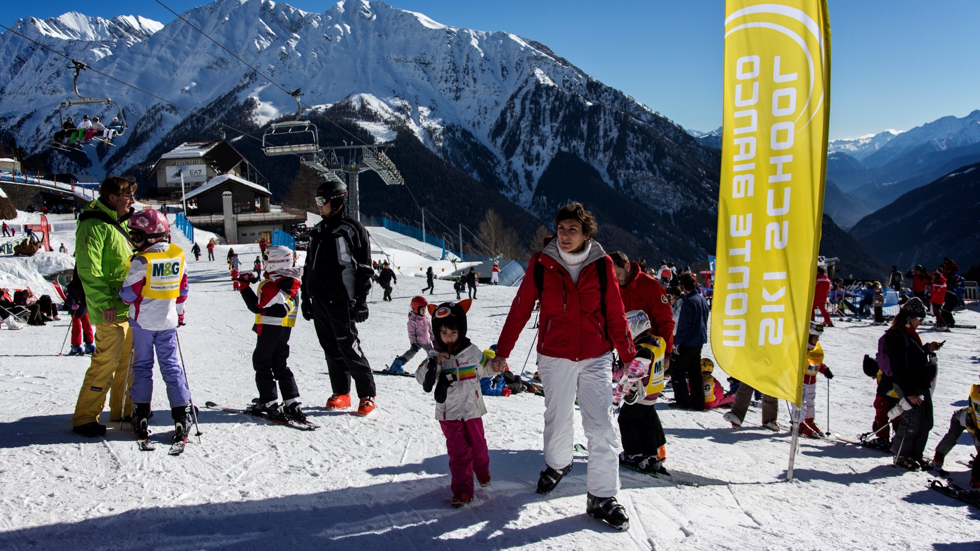 Pista de esqui coberta de neve com esquiadores e snowboarders, um teleférico e montanhas ao fundo.