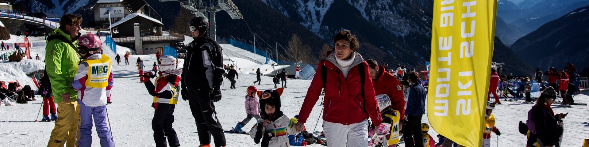 Pista de esqui coberta de neve com esquiadores e snowboarders, um teleférico e montanhas ao fundo.